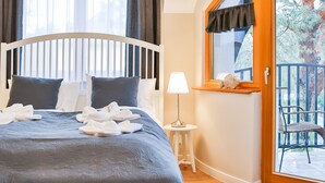 Quarto com uma cama grande, um candelabro, janela com cortinas, uma mesa pequena com luminária e vista para o exterior.