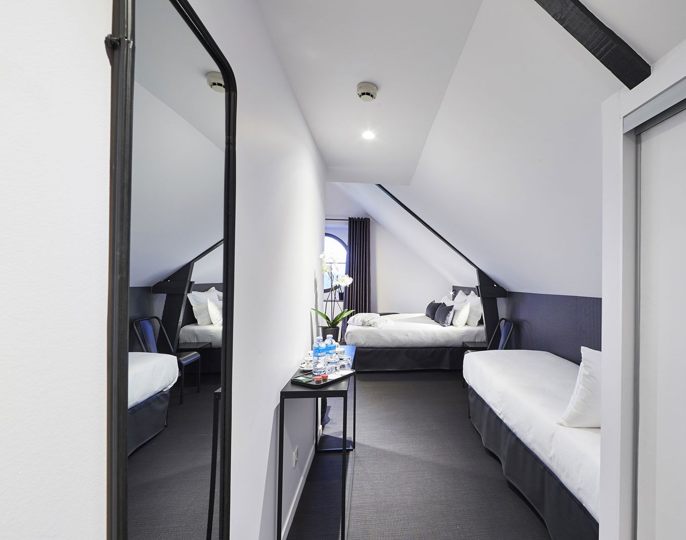 Quarto de hotel moderno com teto inclinado, duas camas individuais, uma mesinha de cabeceira com produtos de higiene e um espelho.