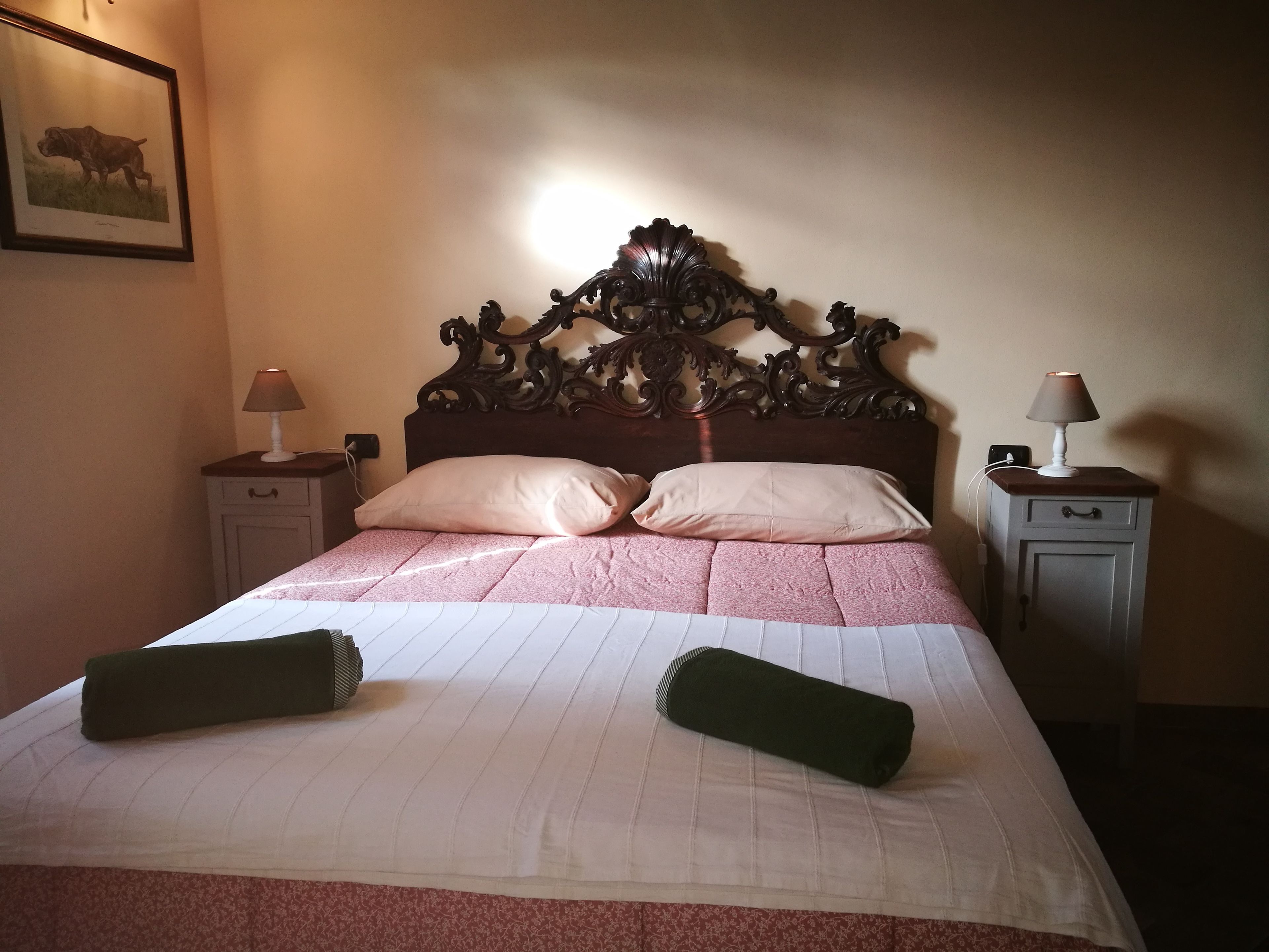 Uma cama bem arrumada com cabeceira decorativa, duas mesas de cabeceira com abajures e um quadro na parede.