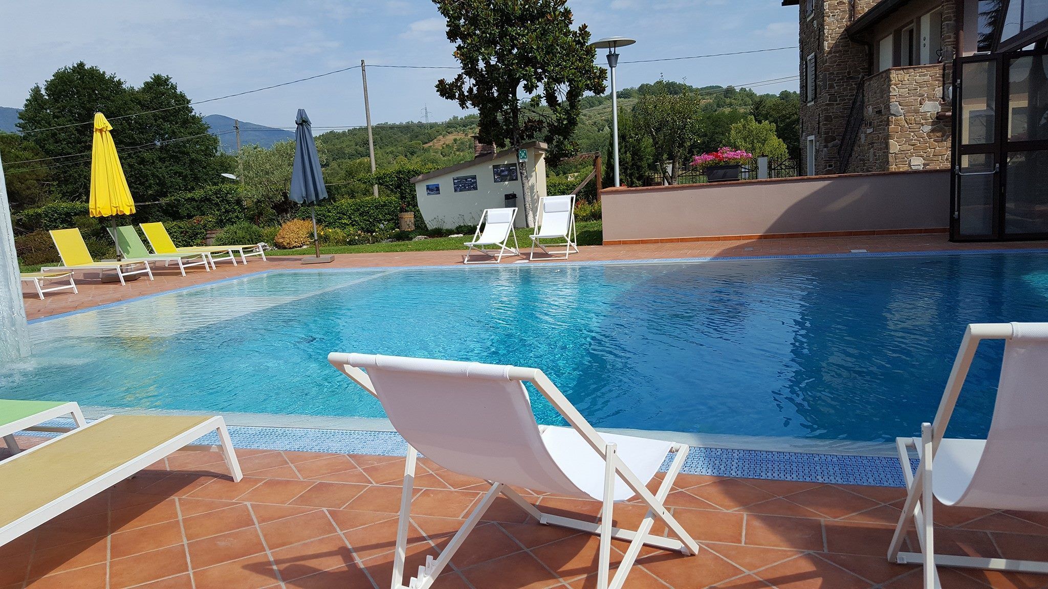 Uma piscina com cadeiras de descanso brancas, um guarda-sol amarelo e um edifício de pedra ao fundo.