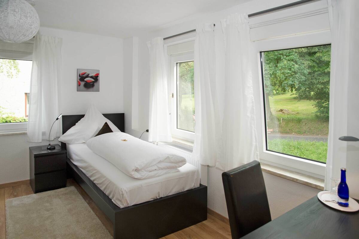 Quarto moderno com cama, mesa de cabeceira, mesa com uma garrafa e vista para o verde lá fora.