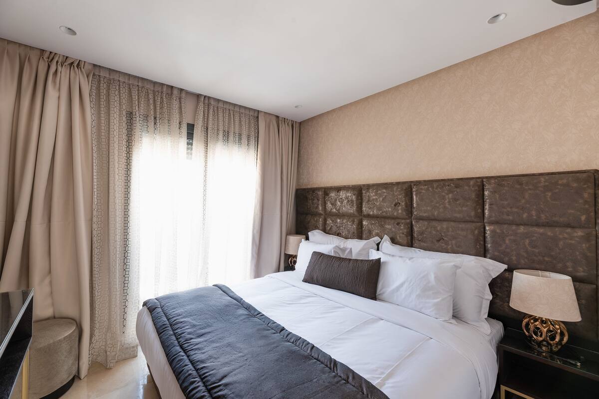 Quarto de hotel com uma cama grande, cabeceira, mesa de cabeceira com abajur e janela com cortinas.