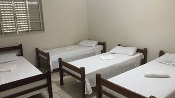 Quarto com quatro camas de solteiro, cada uma com estrutura de madeira e roupa de cama branca. Há uma janela com persianas no lado esquerdo.