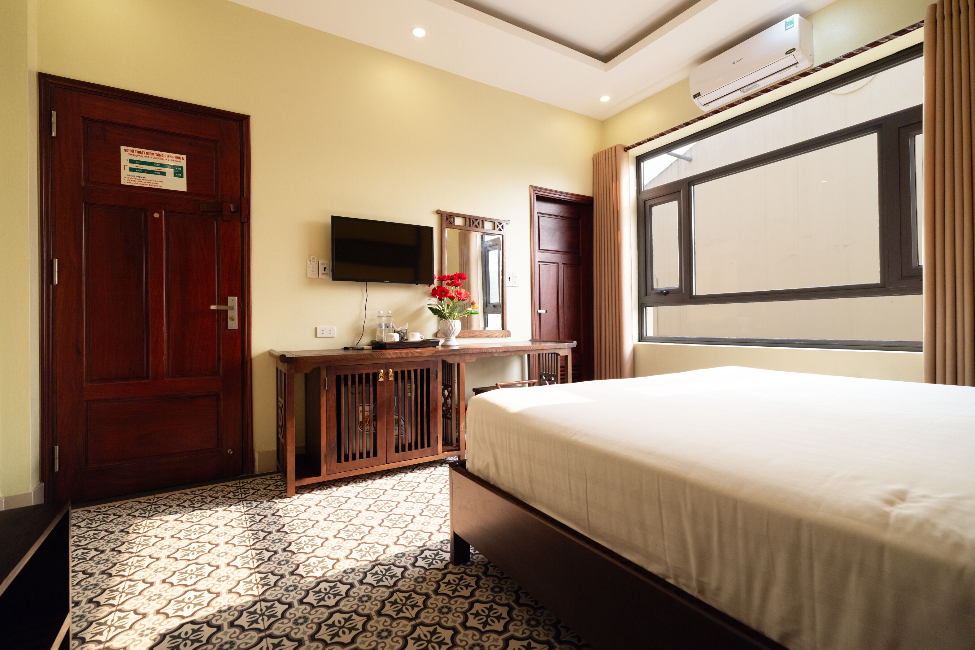 Quarto de hotel com uma cama grande, uma escrivaninha de madeira, uma televisão de tela plana, um espelho e uma janela com cortinas.