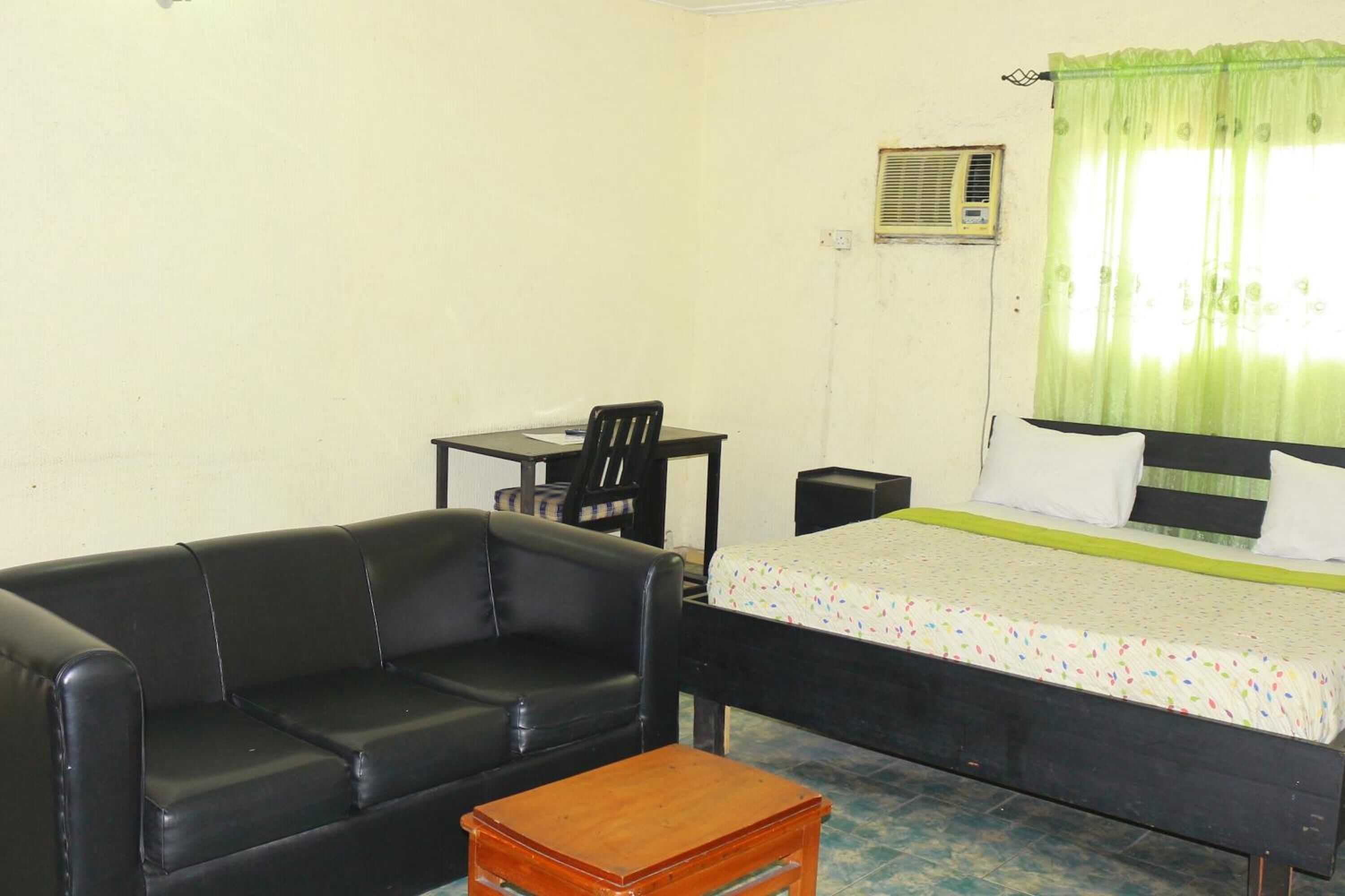 Quarto com sofá de couro preto, mesa de madeira, cama com colcha estampada e cortina verde.