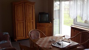 Quarto com armário de madeira, televisão sobre suporte de madeira, mesa de jantar com toalha xadrez e janela com cortinas leves.