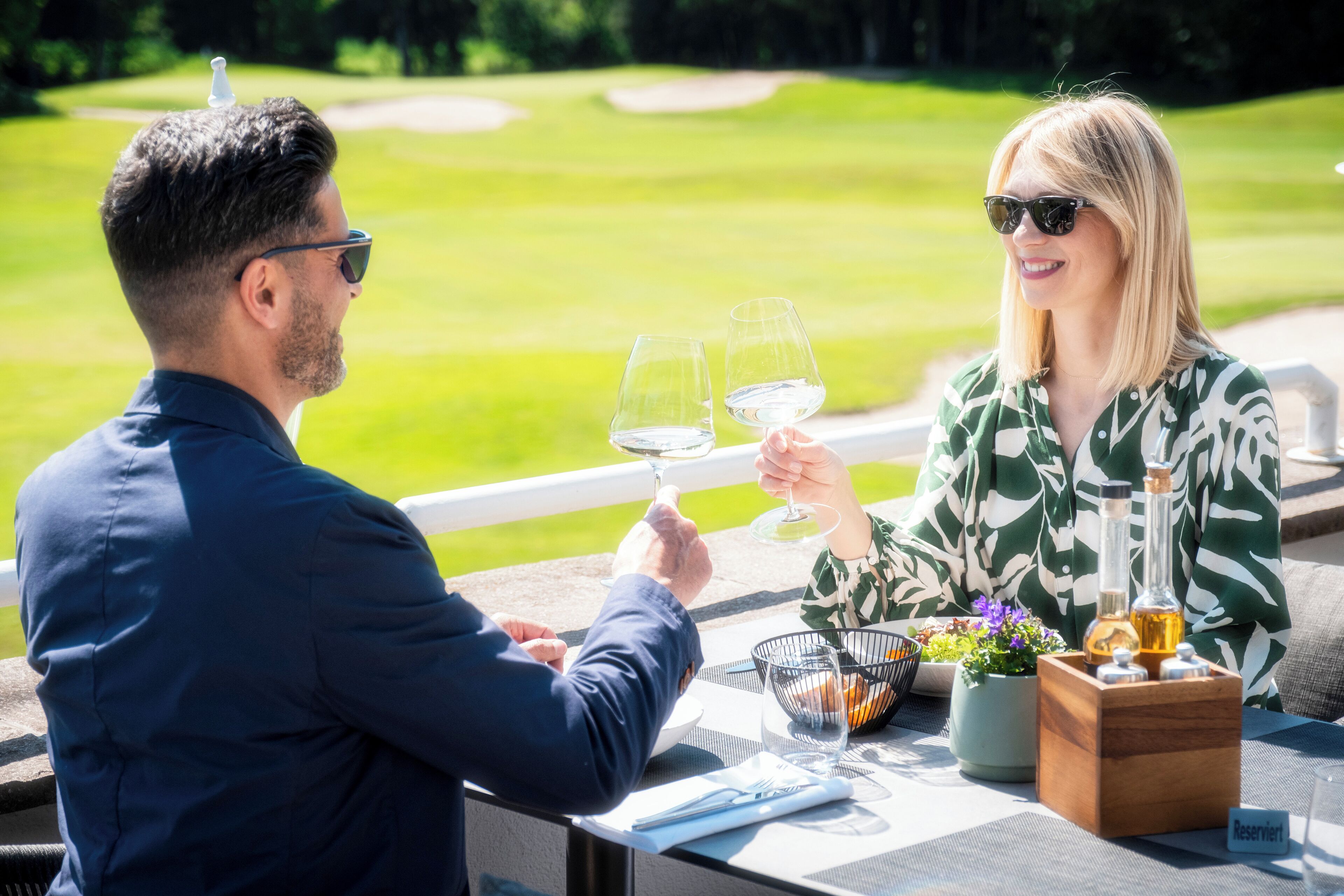Um casal brindando com taças de vinho em uma mesa com vista para um campo de golfe.