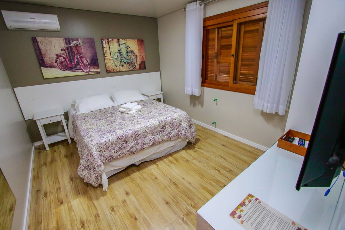 Um quarto com piso de madeira, uma cama com colcha florida, dois quadros fixados na parede retratando bicicletas, uma janela com persianas de madeira e uma escrivaninha com monitor de computador.