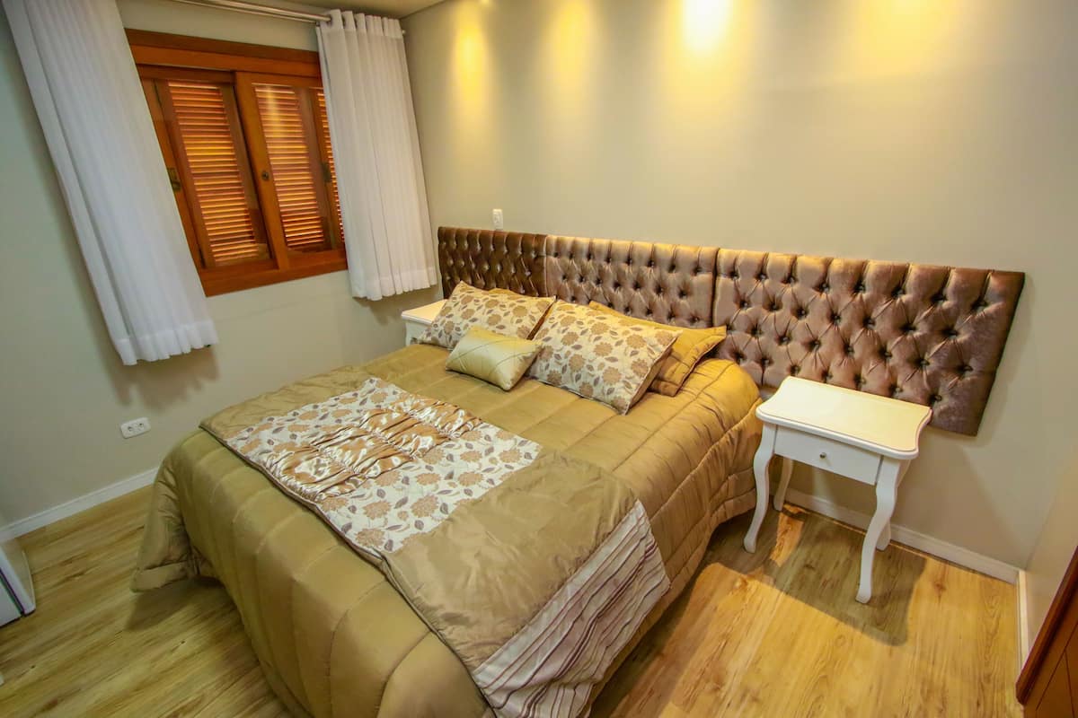 Quarto com uma cama grande, mesa de cabeceira, janela com cortinas e piso de madeira.
