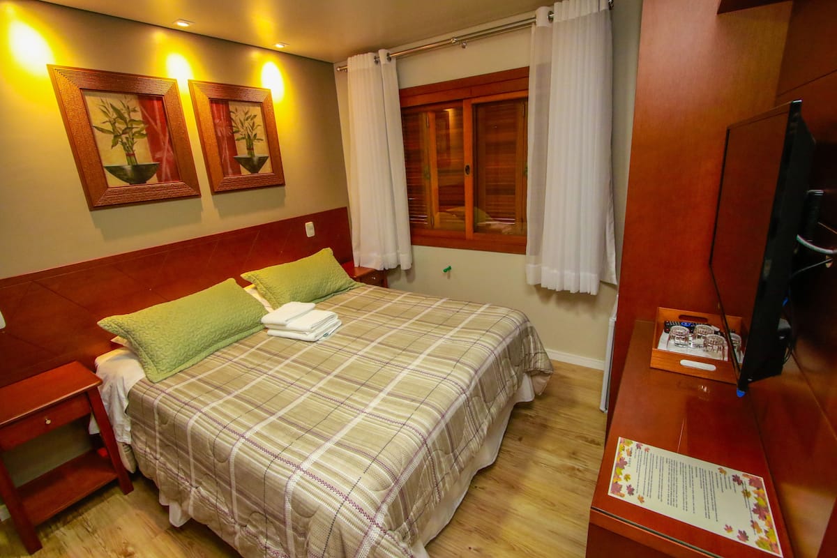 Quarto de hotel com uma cama, mesa de cabeceira, cabeceira de madeira, janela com cortinas e televisão fixada na parede.