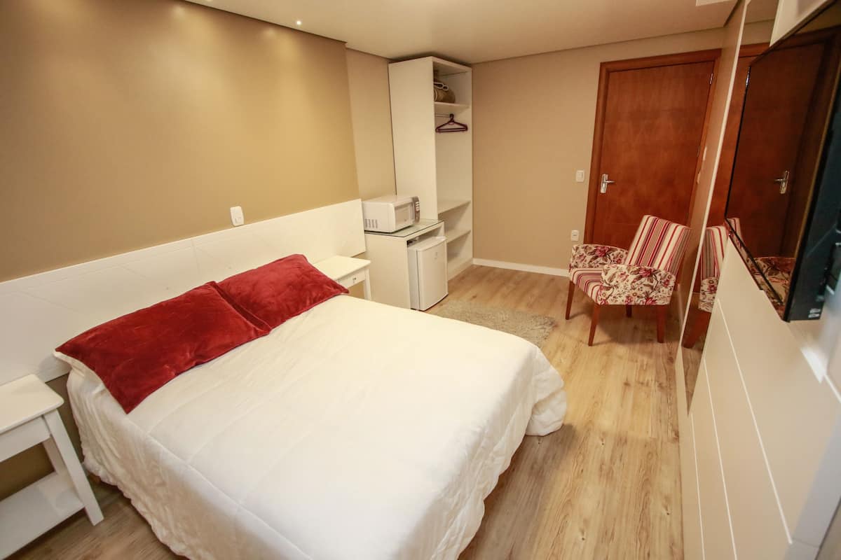 Quarto com cama, mesa de cabeceira, micro-ondas, armário pequeno, cadeira e guarda-roupa.