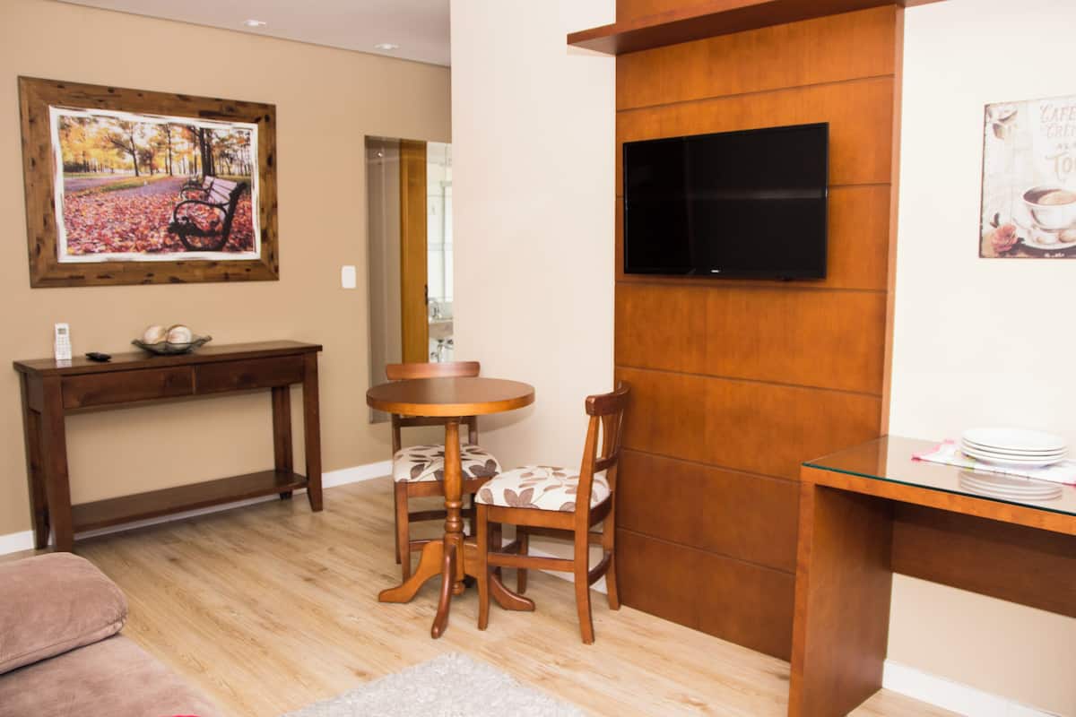 Quarto com escrivaninha de madeira, televisão fixada em um painel de madeira, mesa de jantar com cadeiras e um sofá.