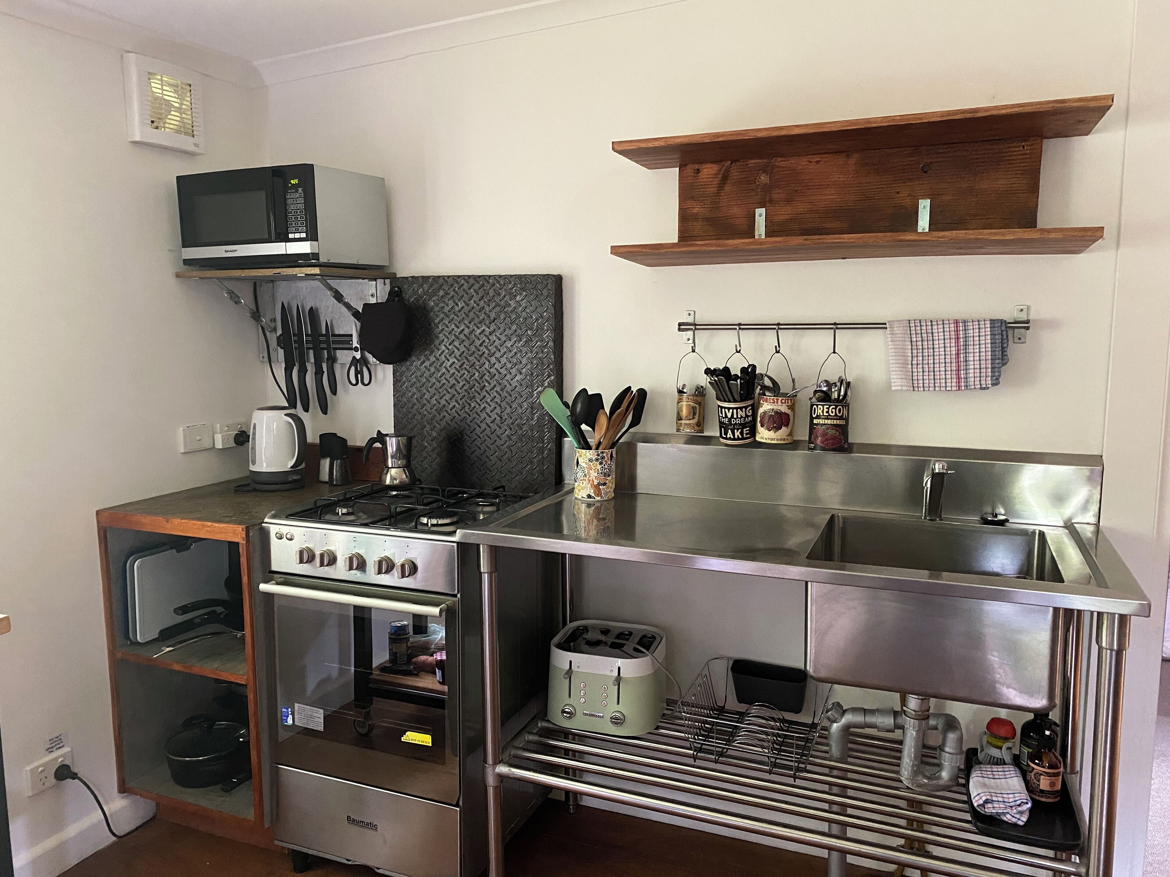 Uma cozinha compacta com fogão, forno, micro-ondas e pia. Há utensílios pendurados na parede e uma prateleira acima da pia.