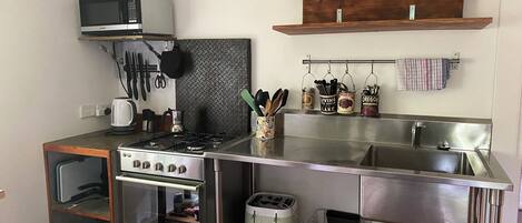 Uma cozinha compacta com fogão, forno, micro-ondas e pia. Há utensílios pendurados na parede e uma prateleira acima da pia.