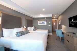 Quarto de hotel com duas camas, uma escrivaninha, uma cadeira, televisão e um quadro na parede.