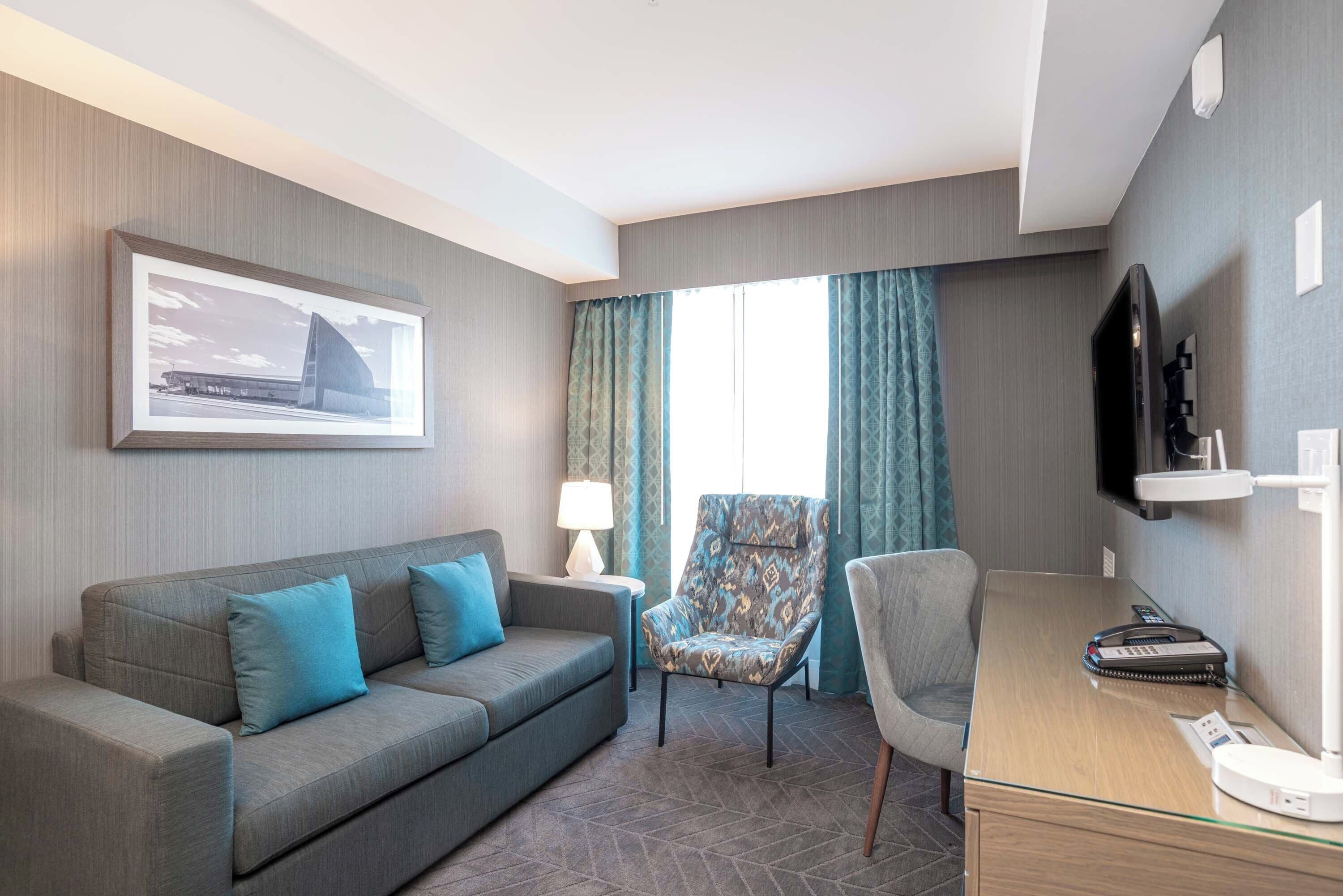 Quarto de hotel com sofá, poltrona, escrivaninha, luminária, TV e um quadro na parede.