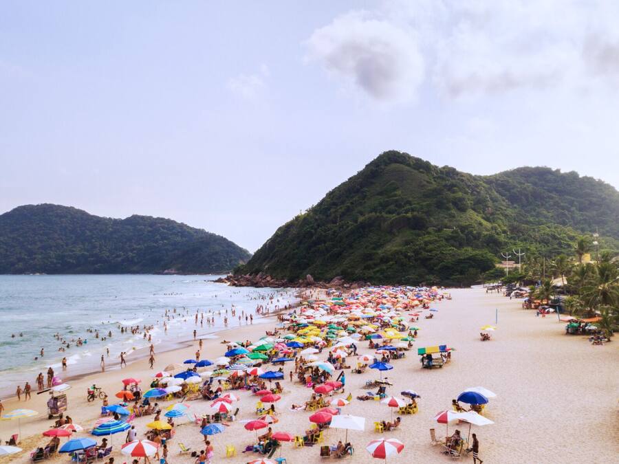 Uma praia cheia, com muitos guarda-sóis coloridos e pessoas aproveitando o mar.