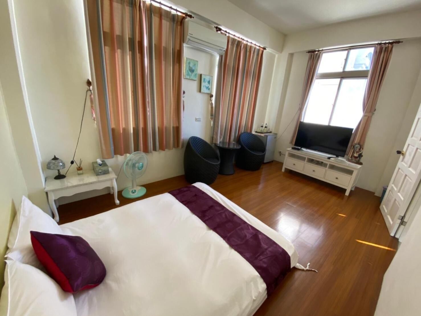 Quarto com cama, escrivaninha, cadeira, televisão e janela com cortinas.