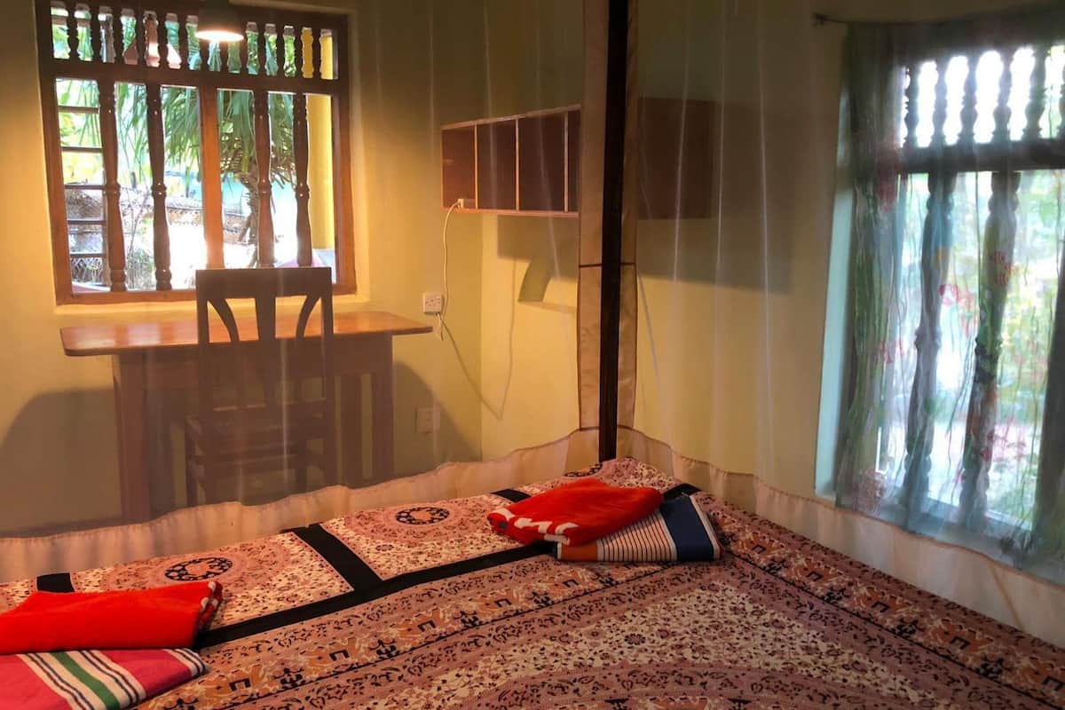 Quarto com cama, escrivaninha e cadeira. Janela com cortinas e vista para o verde lá fora.