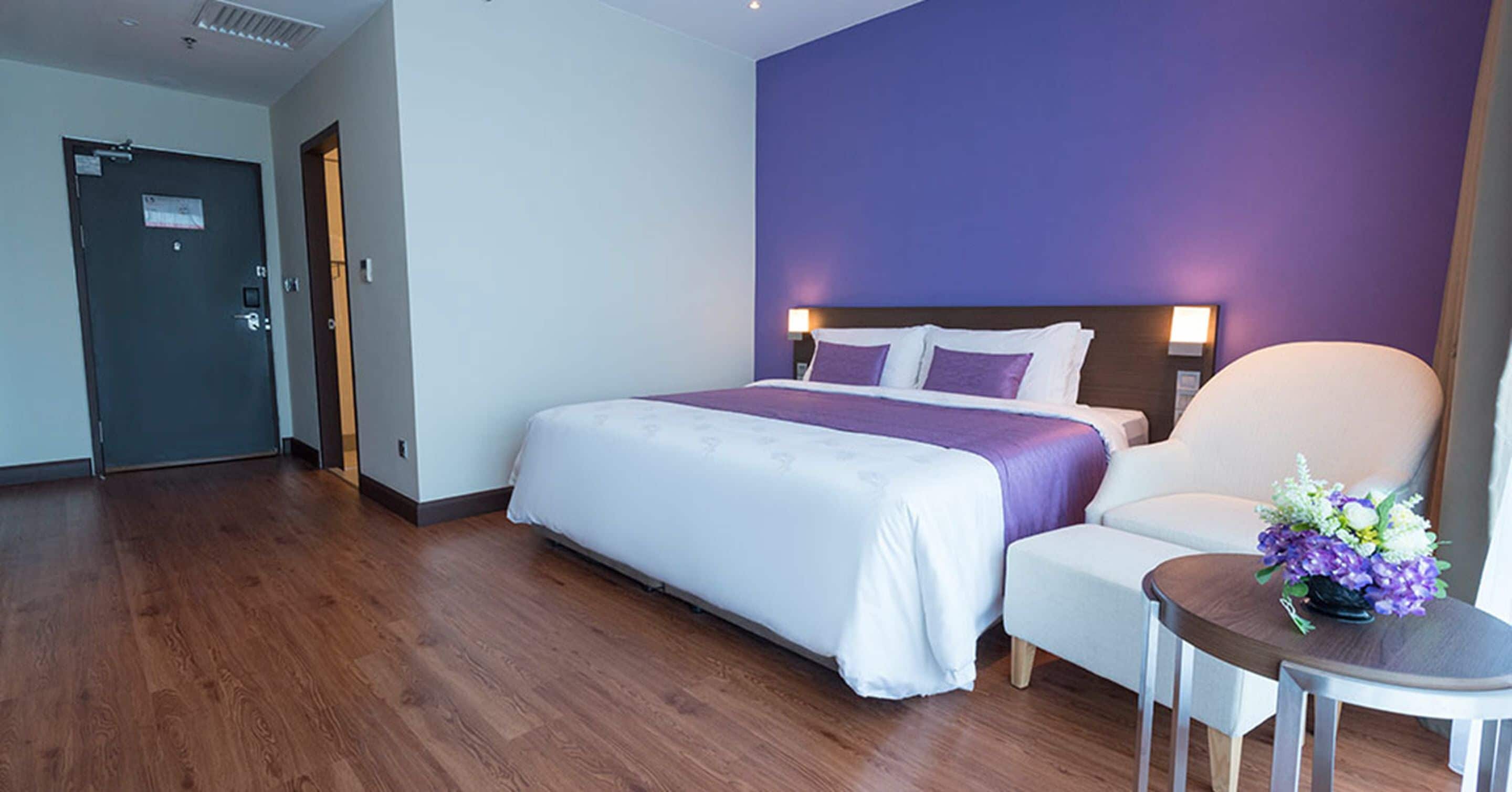 Quarto de hotel com uma cama, uma cadeira e uma pequena mesa com um vaso de flores.