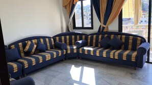 Sala de estar com canto, sofá xadrez azul e amarelo, uma mesinha e vista para prédios através de uma janela com cortinas.