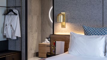Quarto de hotel moderno com uma cama grande, uma mesa de cabeceira com abajur, um roupão pendurado em um porta-roupas e vista para o banheiro.