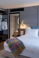Quarto de hotel moderno com uma cama grande, uma mesa de cabeceira com abajur, um roupão pendurado em um porta-roupas e vista para o banheiro.