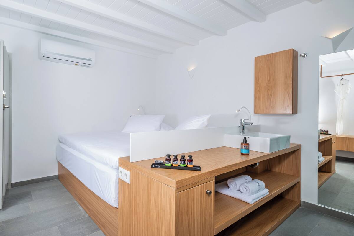 Quarto de hotel moderno com cama, mesa de cabeceira de madeira com artigos de higiene pessoal, pia e espelho.