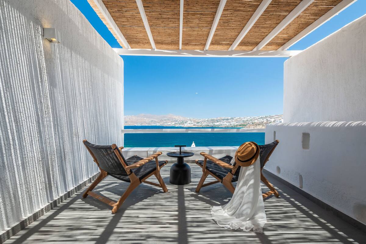 Terraço com vista para o mar, duas cadeiras de vime, uma mesa redonda pequena e um vestido branco no chão.