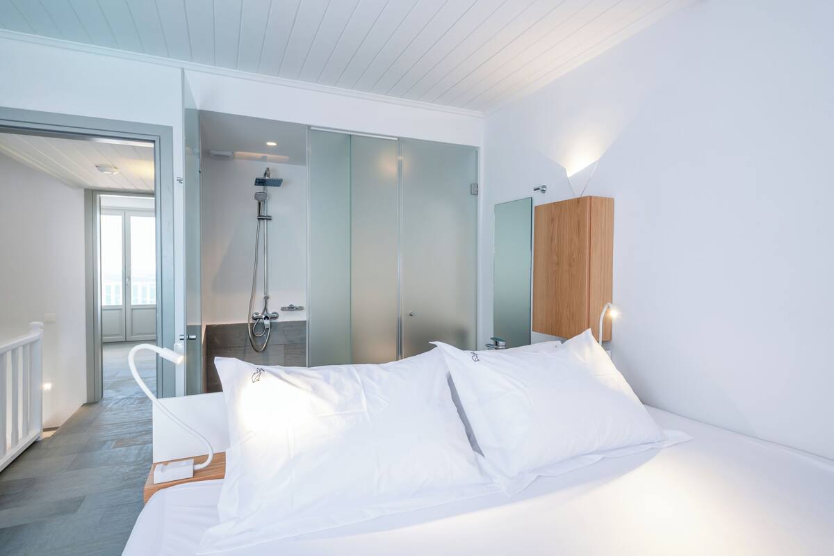 Quarto de hotel moderno com cama, guarda-roupa de madeira, box de chuveiro em vidro e teto branco.