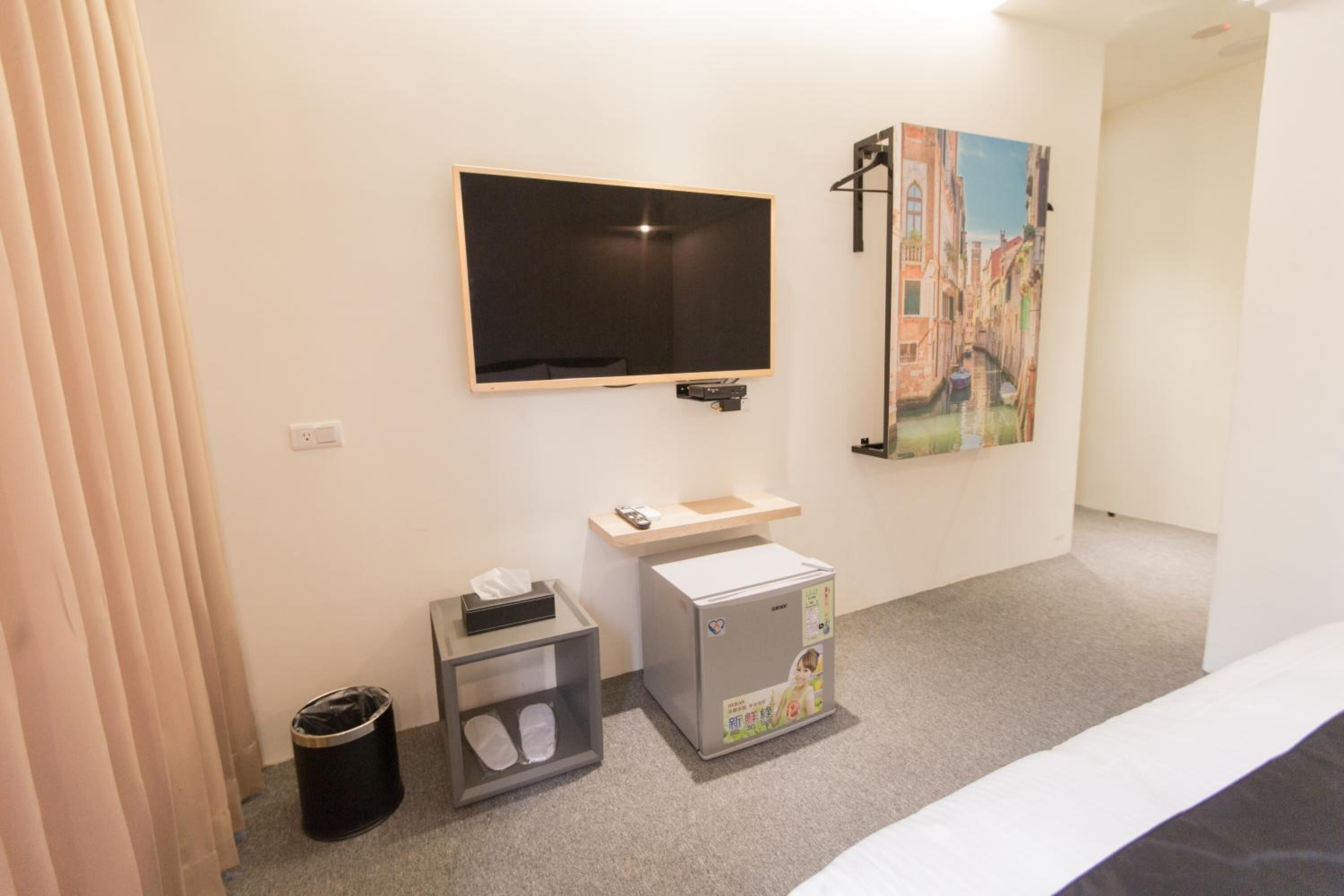Quarto de hotel com televisão de tela plana na parede, mesa de cabeceira pequena com controle remoto, lixeira e mini-geladeira.