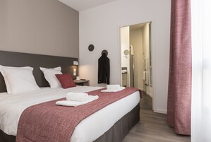 Quarto de hotel com uma cama grande, uma mesa de cabeceira com abajur e um banheiro visível através de uma porta aberta.