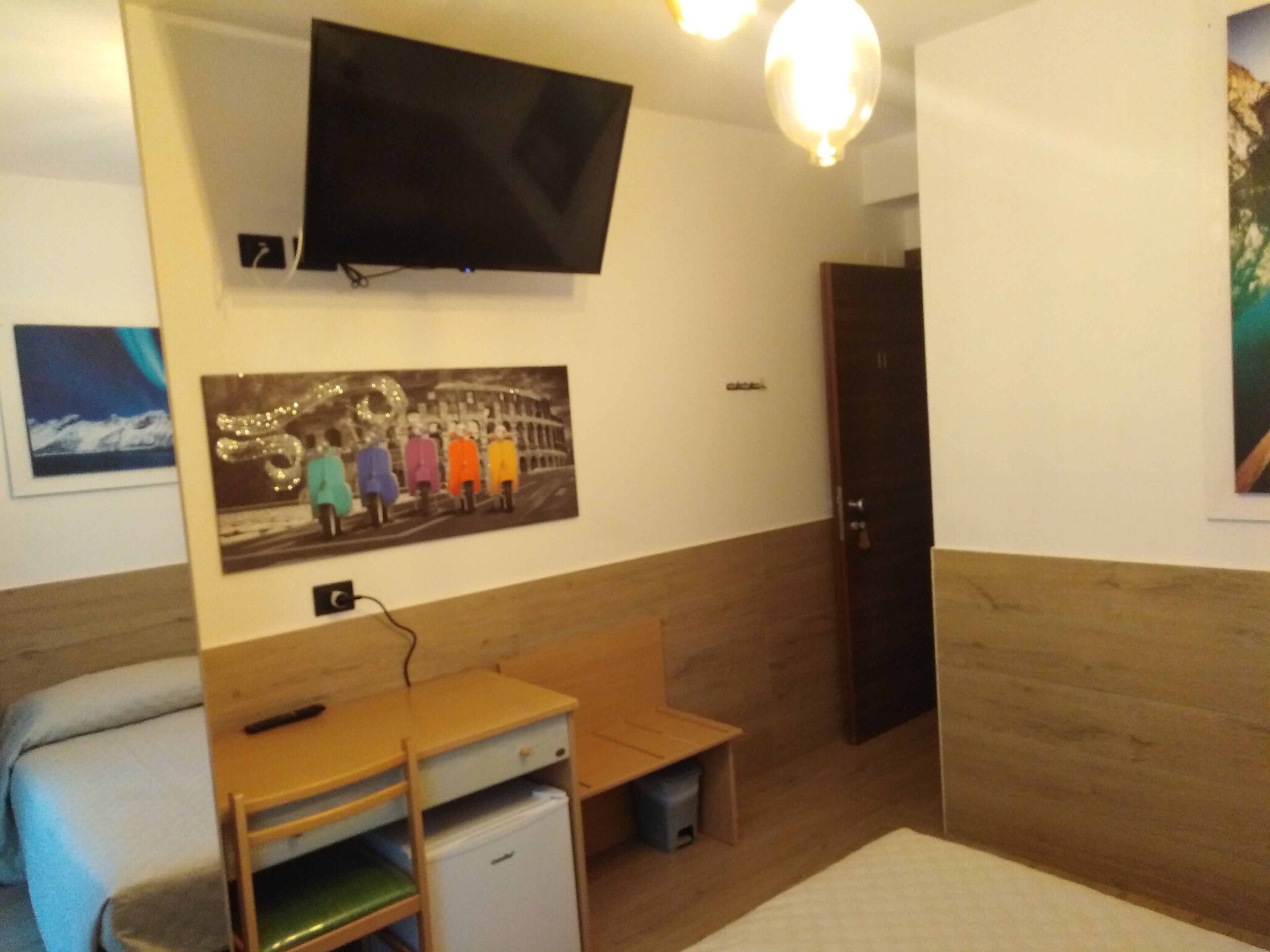 Quarto com televisão de tela plana na parede, um banco de madeira, uma pequena geladeira e uma porta.