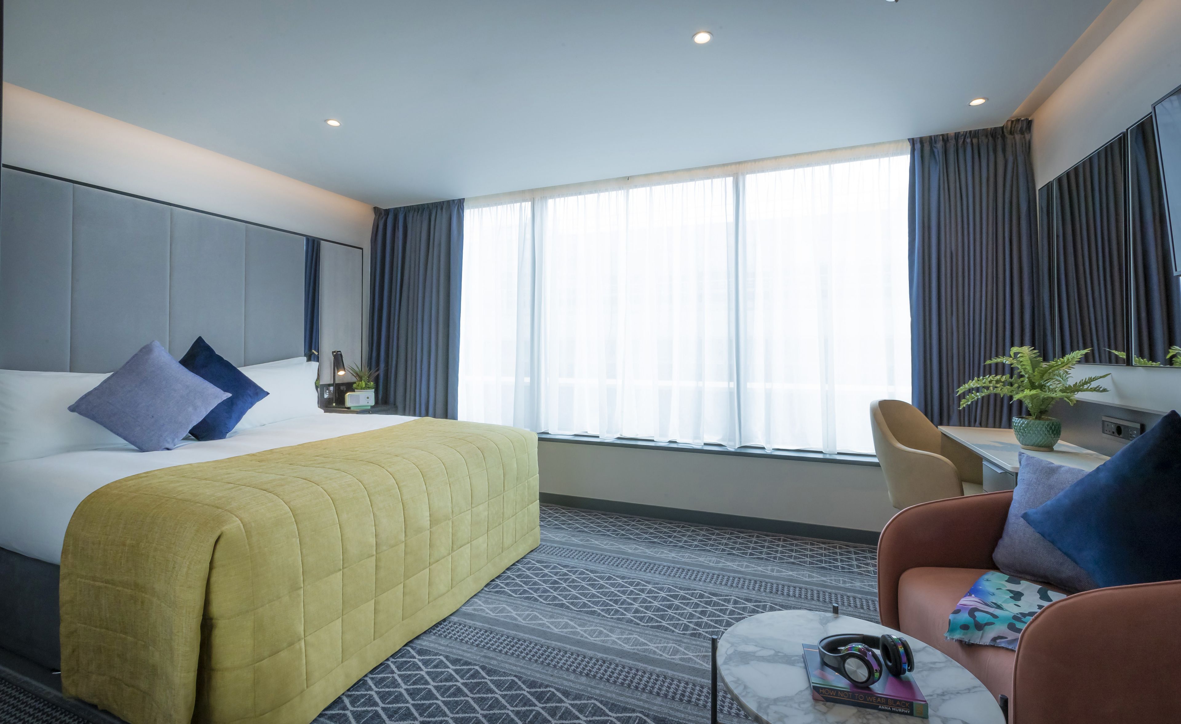 Quarto de hotel com uma cama grande, uma escrivaninha com cadeira, uma mesa pequena e uma janela ampla com cortinas.