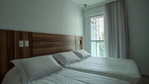 Quarto de hotel com duas camas, cabeceira de madeira, roupa de cama branca, janela com cortinas e vista para edifícios.