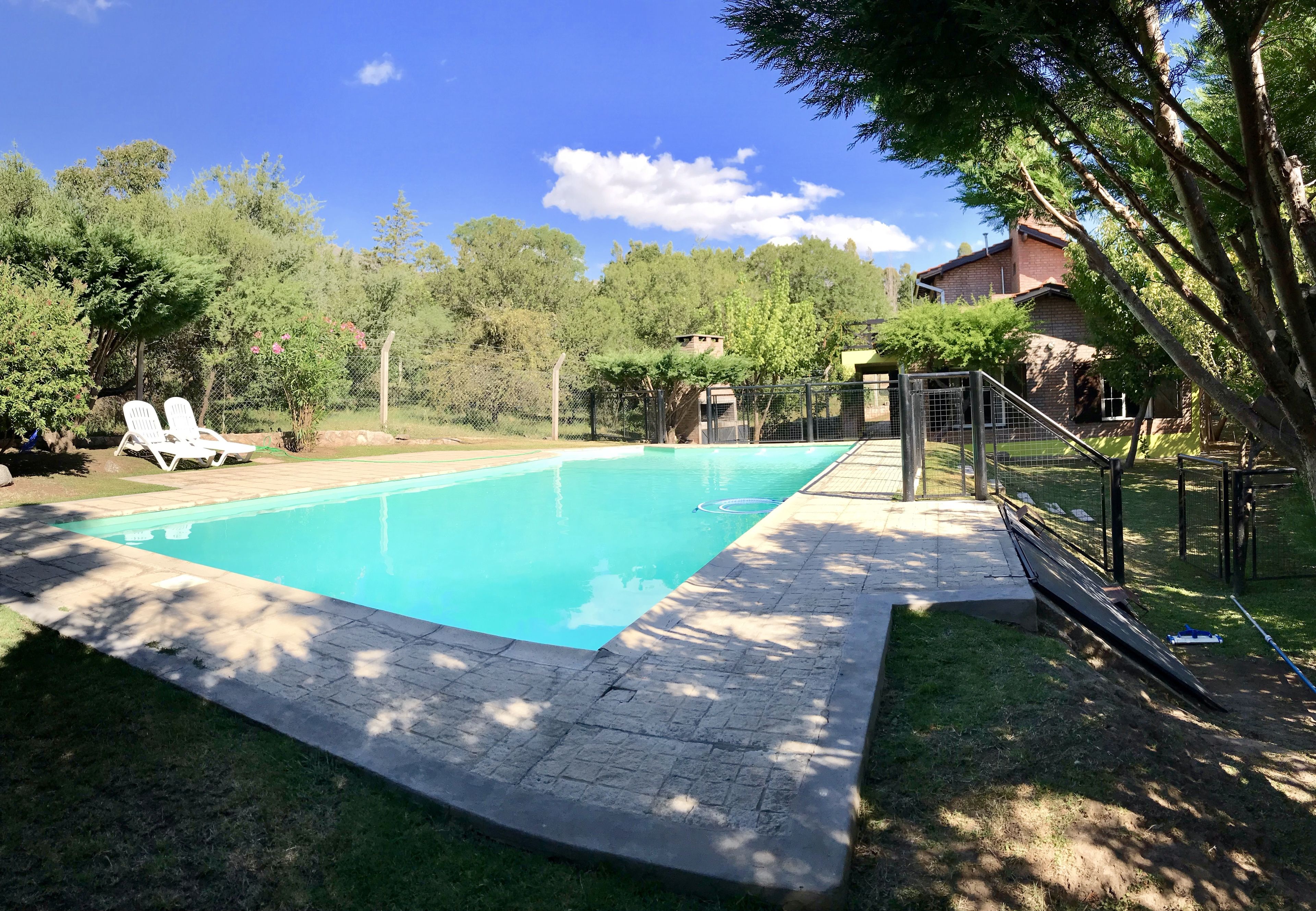 Piscina retangular com deck pavimentado, cercada por uma vedação e árvores.