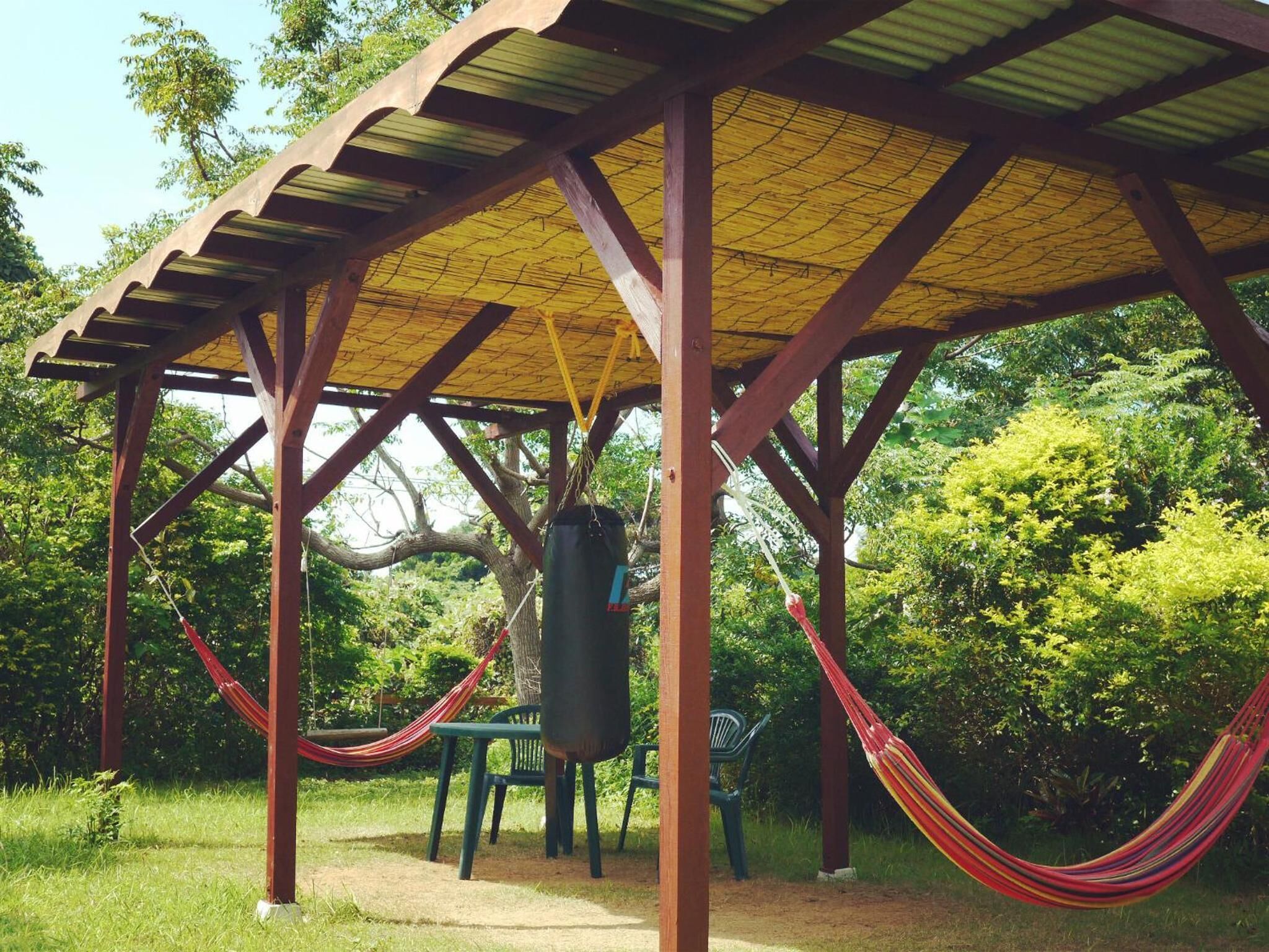 Um gazebo de madeira com telhado de palha, uma rede e duas cadeiras em um jardim exuberante.