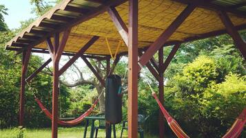 Um gazebo de madeira com telhado de palha, uma rede e duas cadeiras em um jardim exuberante.