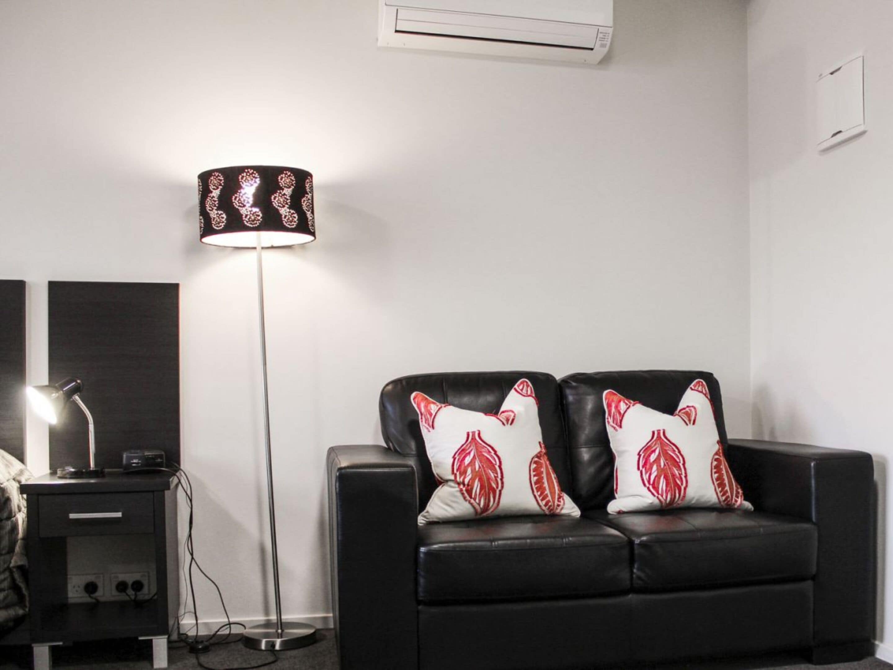 Uma sala de estar moderna com um sofá de couro preto, duas almofadas decorativas, um abajur, uma mesinha lateral e um aparelho de ar condicionado na parede.