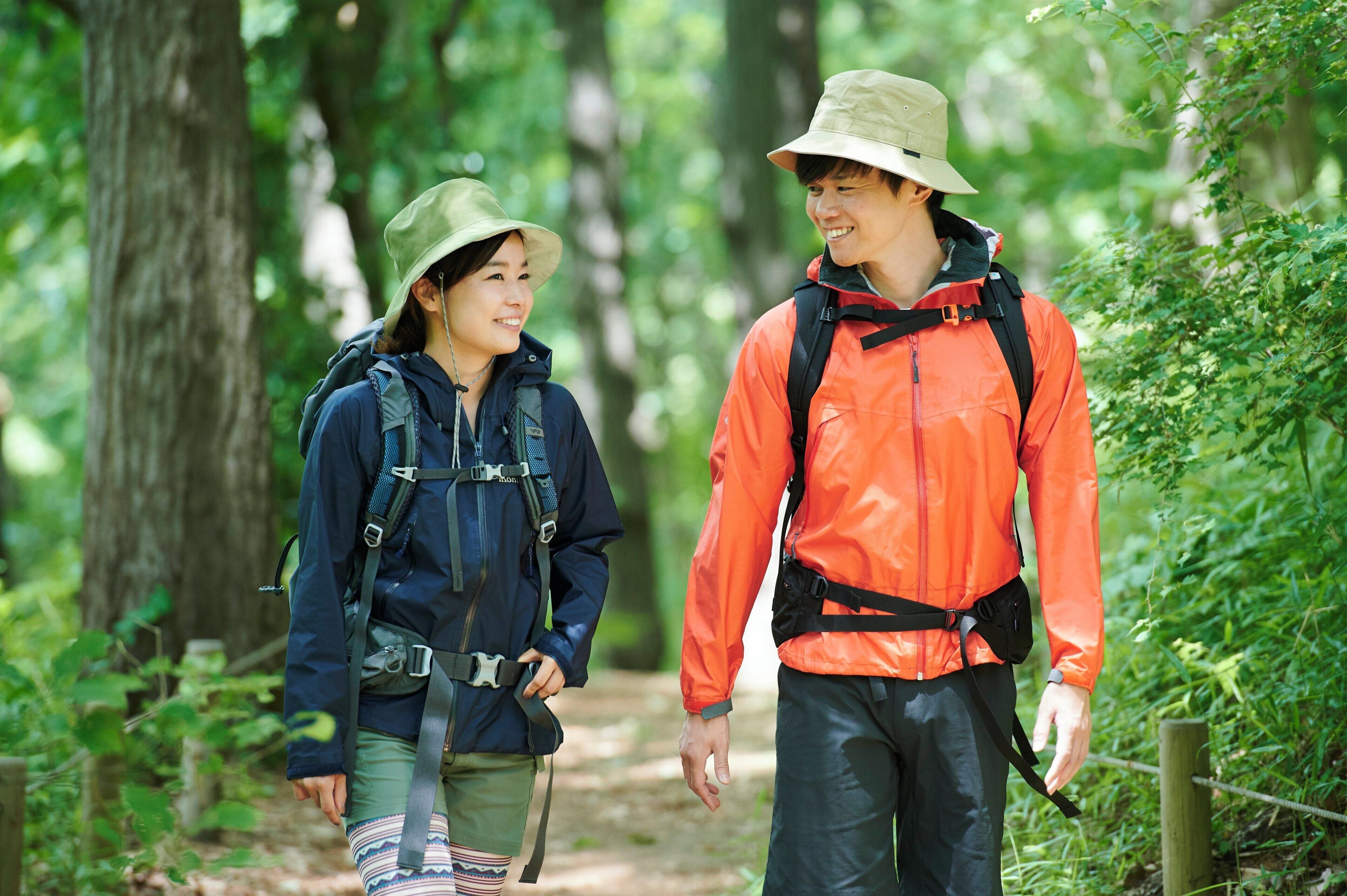 Duas pessoas caminham por uma trilha na floresta, uma vestindo jaqueta azul e calça verde, a outra com jaqueta laranja e calça preta.