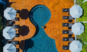 Vista aérea de uma piscina com formato peculiar, cercada por espreguiçadeiras e guarda-sóis.