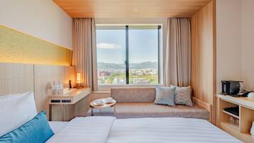 Quarto de hotel com uma cama grande, área de estar com almofadas, uma mesa pequena com xícara e pires, e vista para a paisagem urbana através de uma janela ampla.