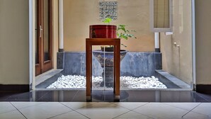 Um corredor com piso ladrilhado, uma mesa de madeira com planta em vaso, um objeto decorativo na parede e uma janela com persianas.