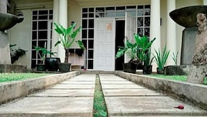 Entrada de edifício com caminho central, colunas e plantas em vasos.