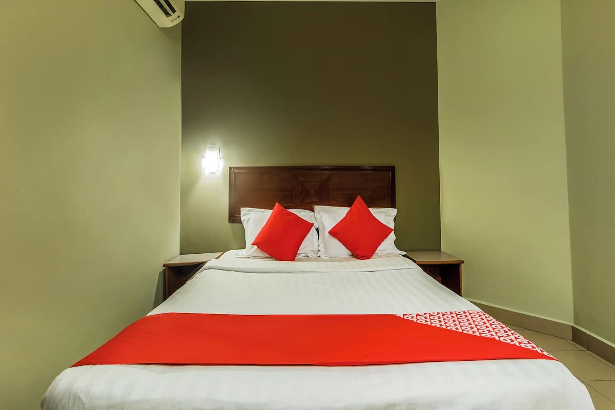 Uma cama bem arrumada com roupa de cama branca e detalhes vermelhos, uma cabeceira de madeira e uma pequena mesa de cabeceira com abajur.