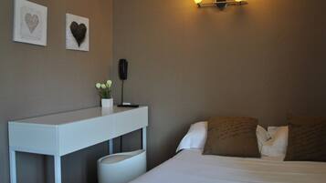 Quarto de hotel com cama, escrivaninha, telefone e duas luminárias fixadas na parede.