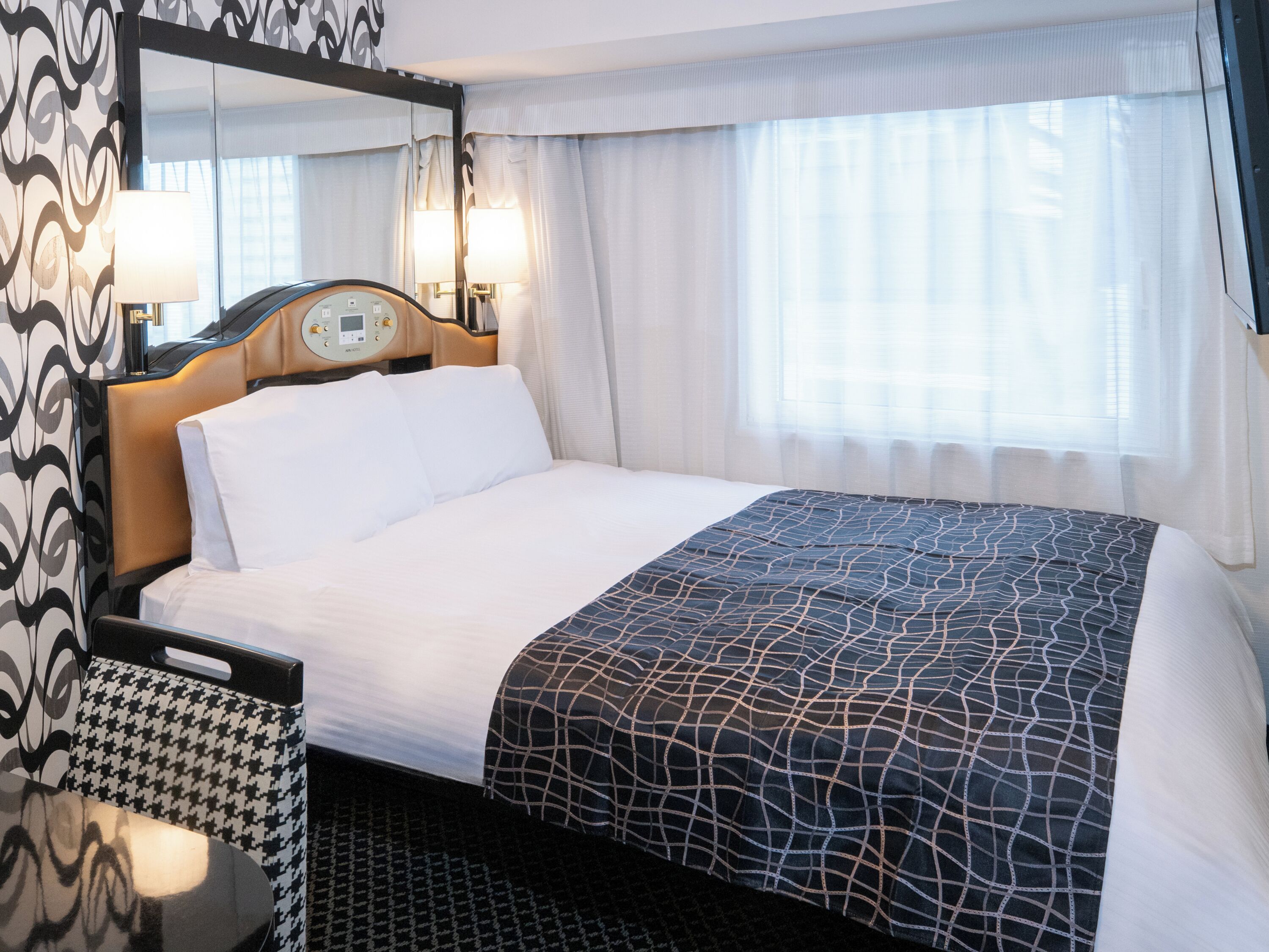Quarto de hotel com cama, mesa de cabeceira, luminária, espelho e janela com cortinas.