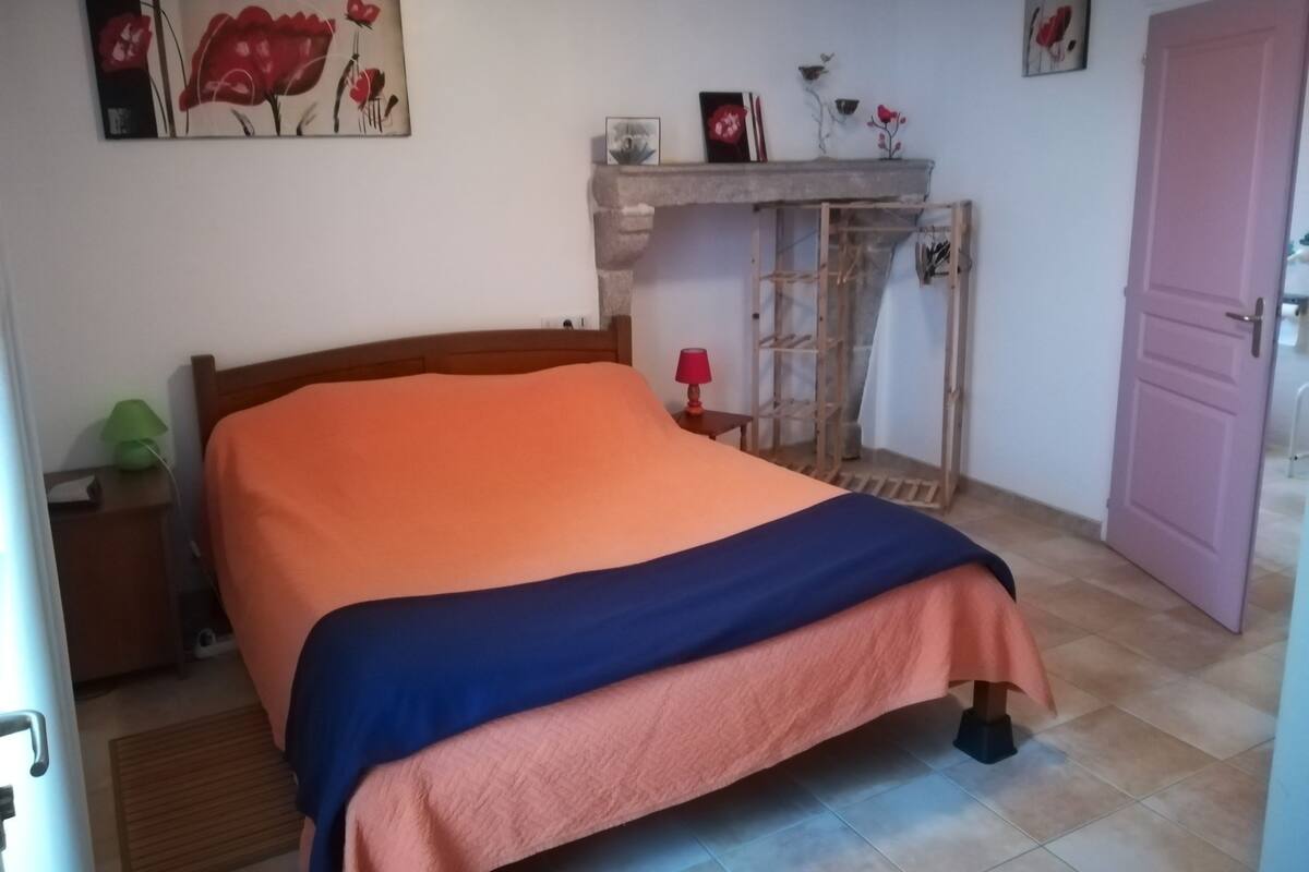 Quarto com cama, mesa de cabeceira, cômoda, prateleira de madeira com objetos decorativos, porta rosa e parede com arte floral.