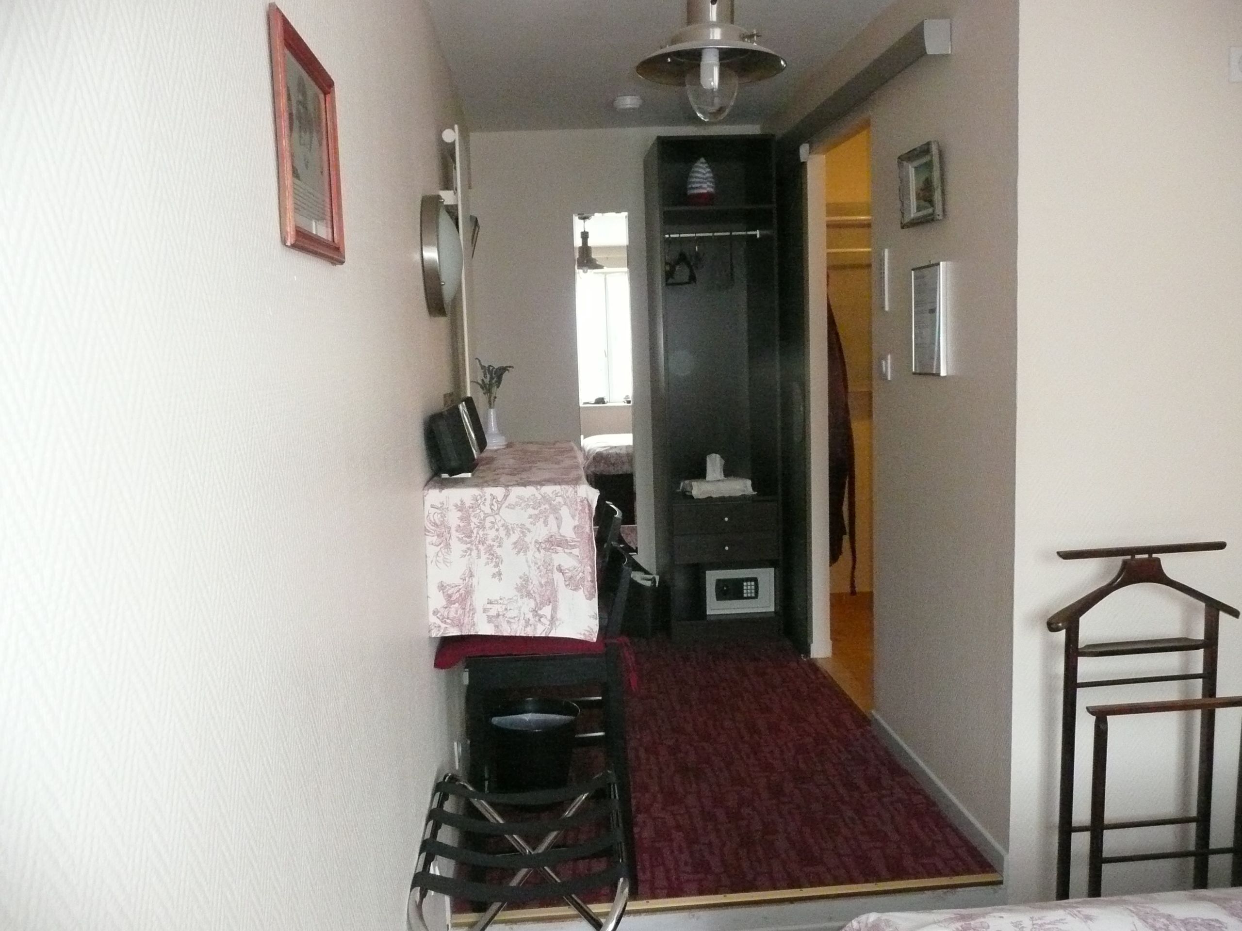 Um corredor estreito com uma cama no final, uma pequena mesa com toalha de mesa florida, um tapete vermelho, um cabideiro e uma porta que dá para outro quarto.