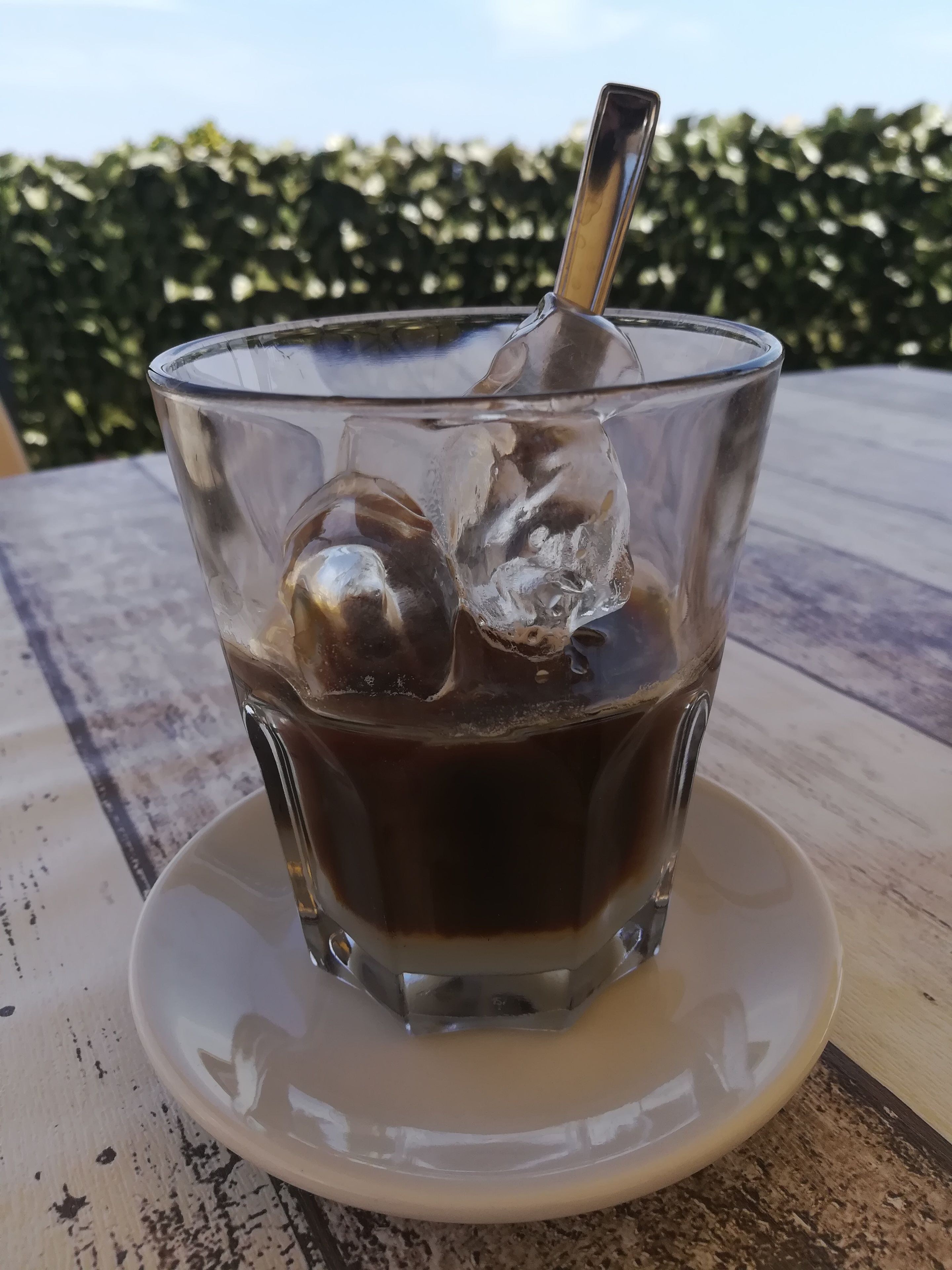 Uma taça de vidro com uma colher, contendo um líquido escuro e cubos de gelo, apoiada num pires.